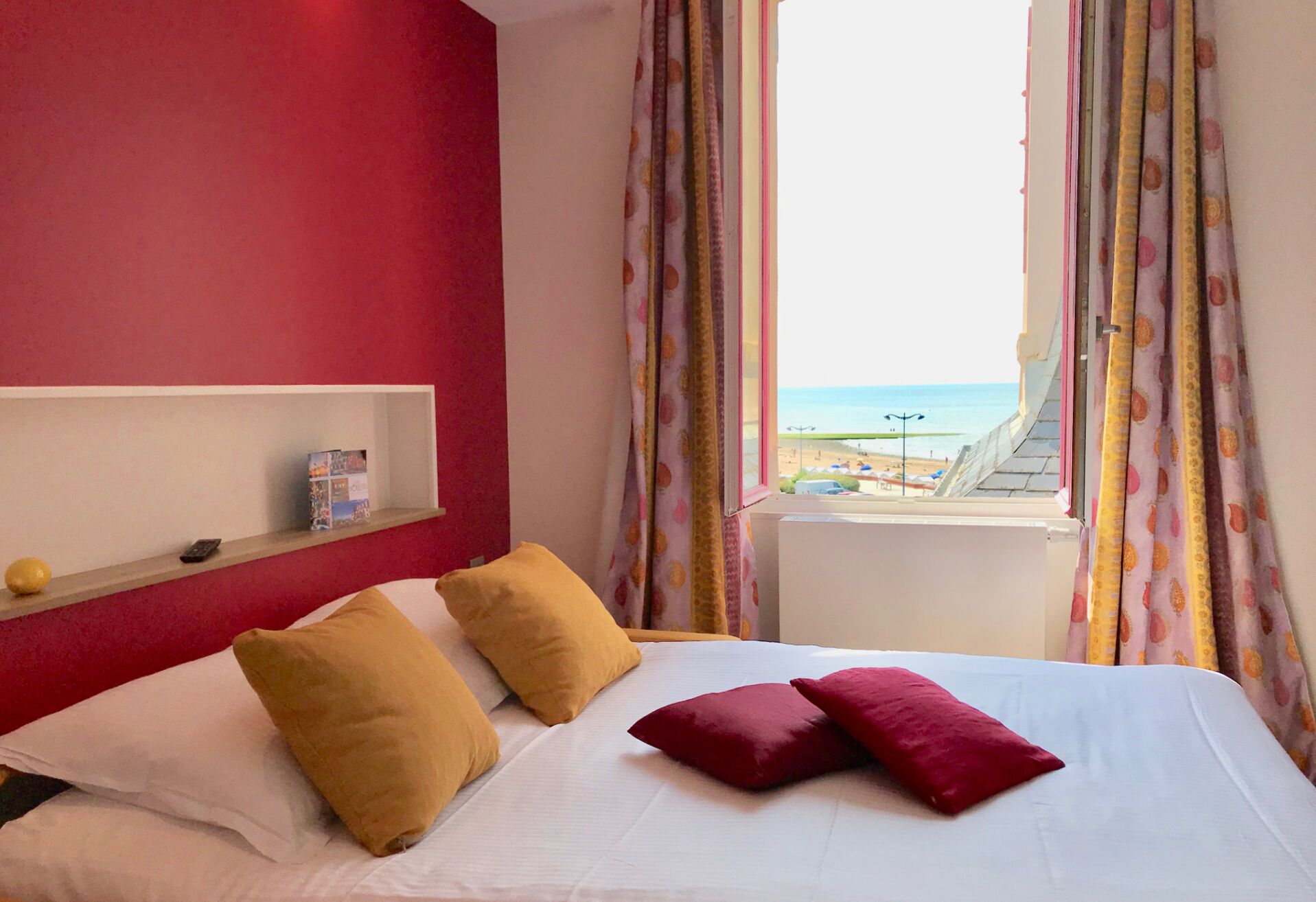 Quarto de hotel com uma cama, duas almofadas, uma prateleira com objetos e vista para a praia.