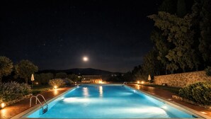 Piscina retangular à noite, com deck de madeira, rodeada por árvores e um muro de pedra.