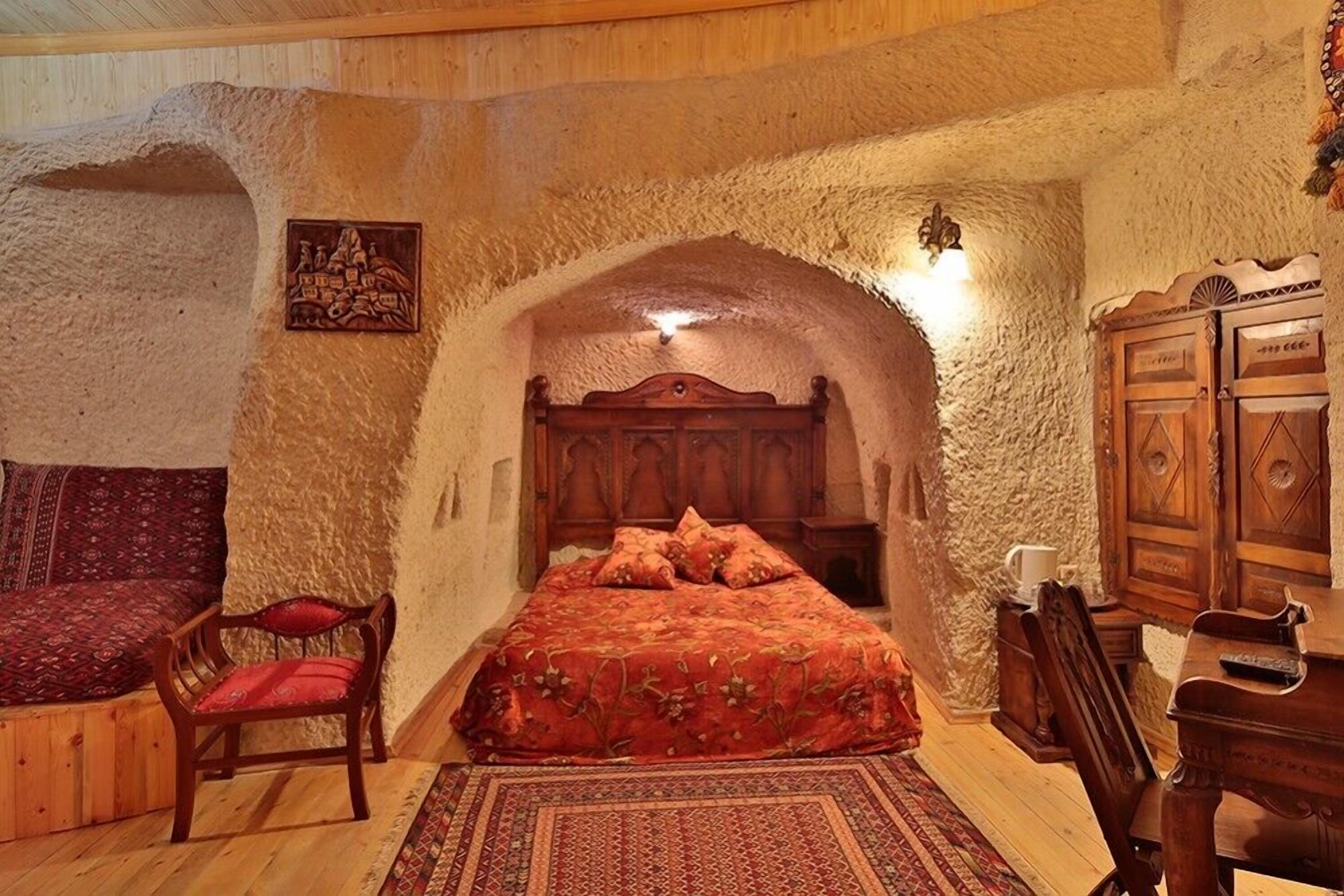 Um quarto aconchegante com parede de pedra, cama de madeira com roupa de cama vermelha, uma mesa pequena e uma cadeira.