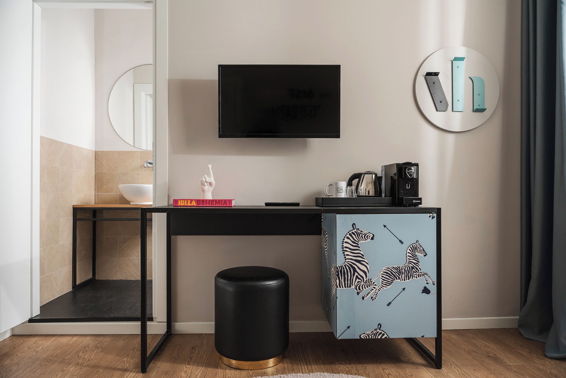 Quarto de hotel moderno com uma escrivaninha preta, televisão de tela plana fixada na parede, um pequeno armário com padrão de zebra, lixeira preta e espelho redondo.