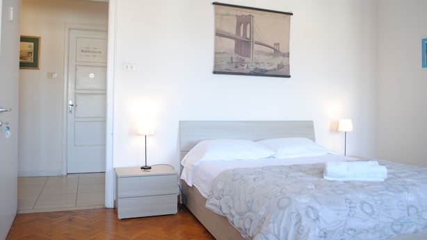 Quarto com cama, luminárias de cabeceira, mesa de cabeceira, porta e quadro pendurado na parede retratando uma ponte.