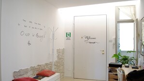 Quarto de hotel com parede de destaque em pedra, um banco branco com almofadas vermelhas, uma escrivaninha com uma planta e uma porta com placa de boas-vindas.