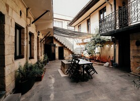 Uma viela estreita com uma mesa e cadeiras no centro, com edifícios laterais com varandas e plantas em vasos.