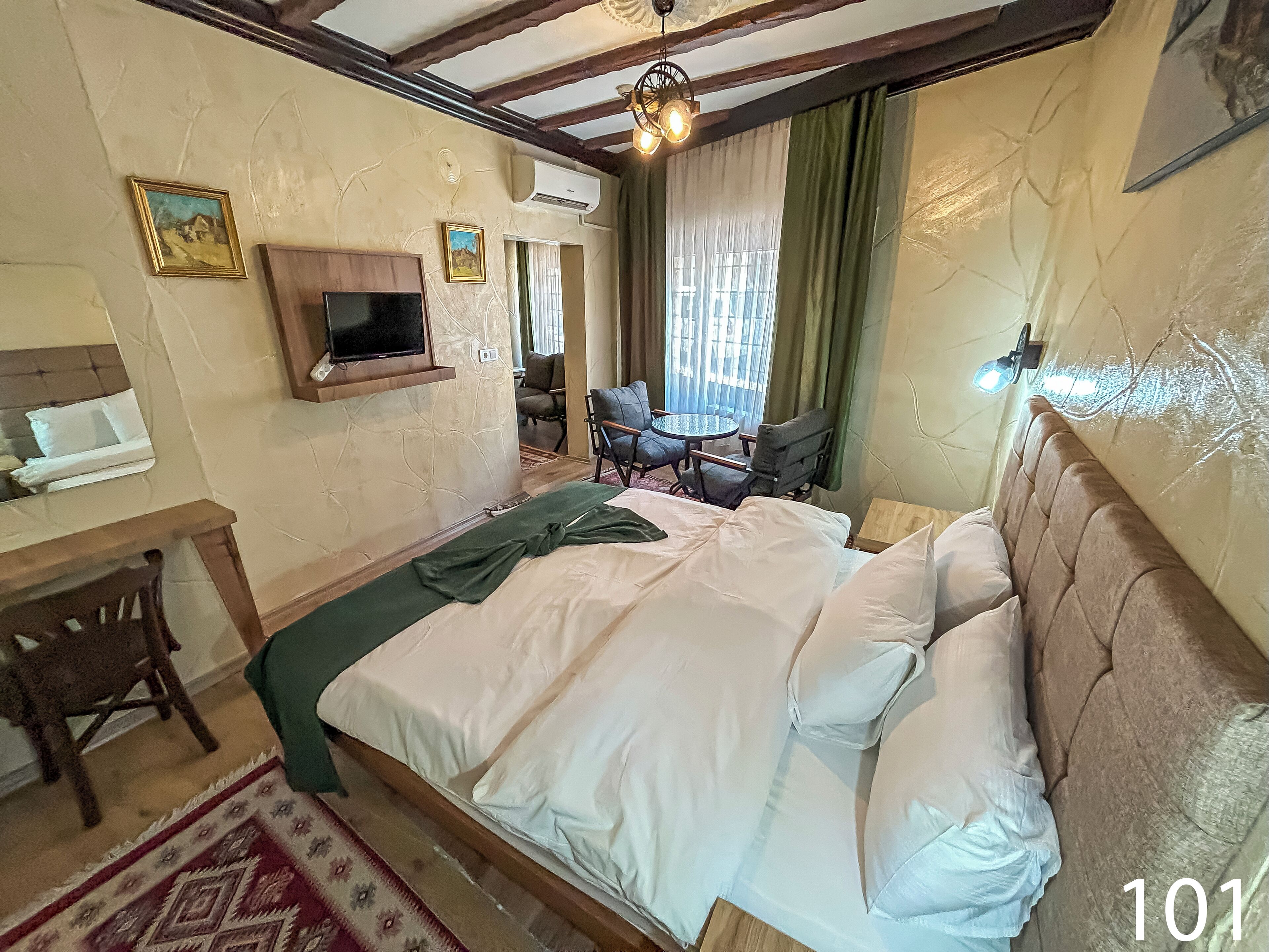 Quarto com cama, escrivaninha, cadeira, televisão e janela com cortinas.