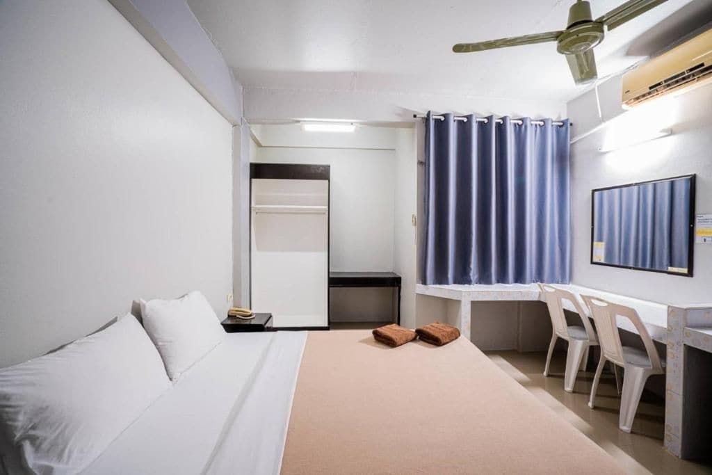 Quarto de hotel com cama, escrivaninha, cadeira, ventilador de teto e janela com cortinas.