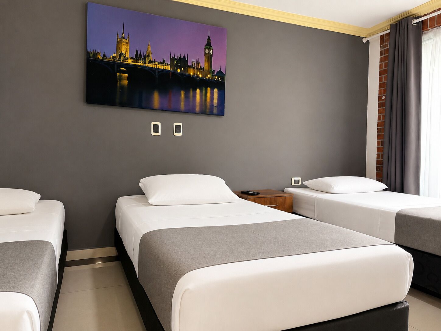 Quarto de hotel com duas camas, uma mesinha de cabeceira e um quadro na parede retratando uma paisagem urbana.