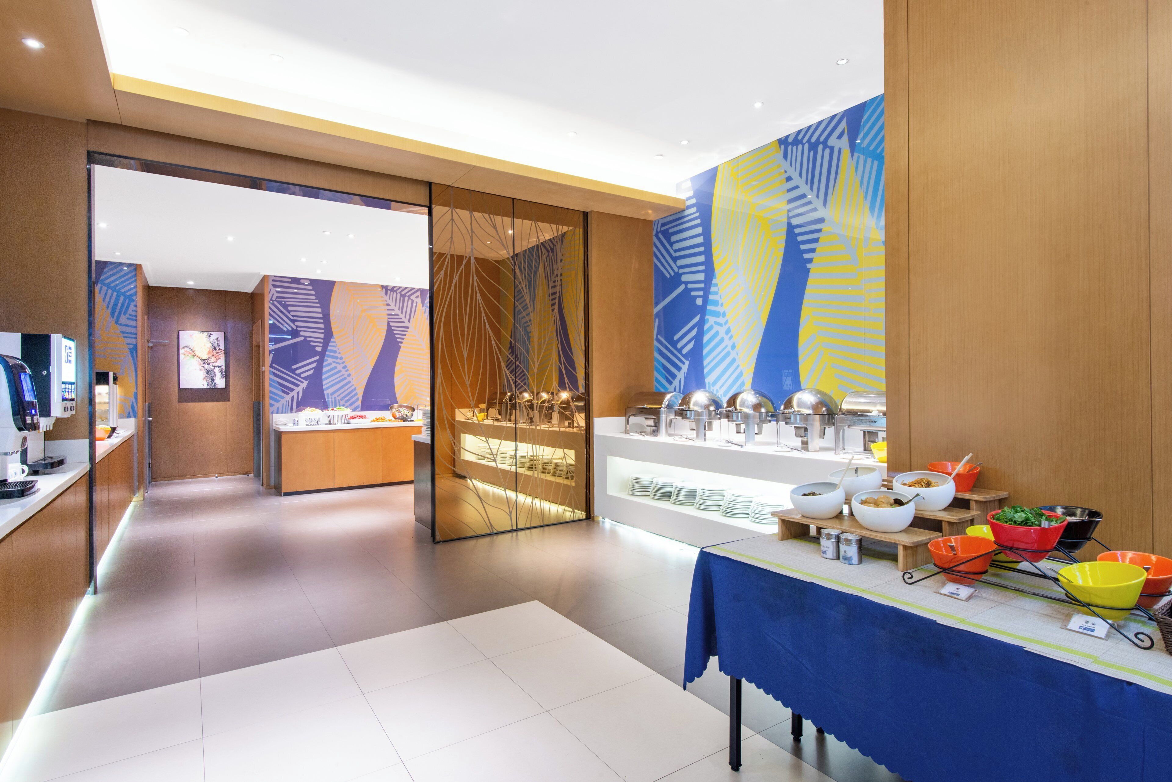 Um hall de hotel moderno com balcão de recepção, área de buffet com diversos alimentos e um espaço de estar com toalha de mesa azul.