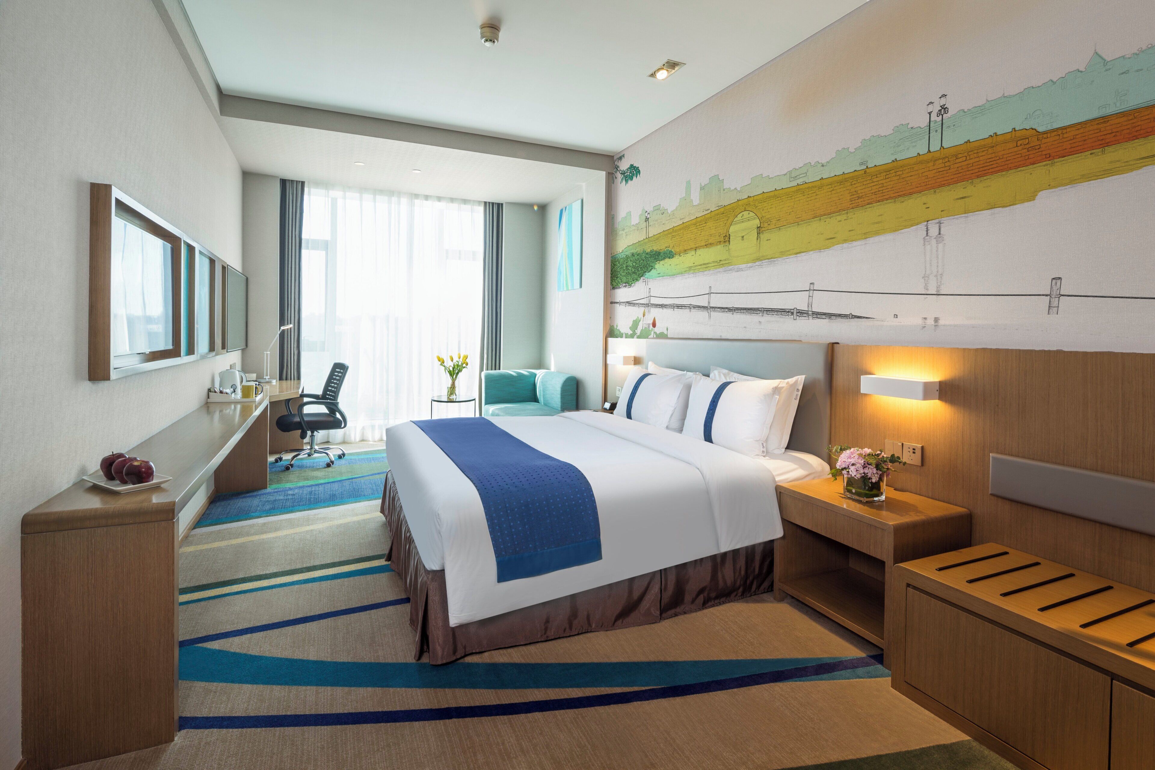 Quarto de hotel com cama, escrivaninha, cadeira, mesa de cabeceira e televisão fixada na parede.