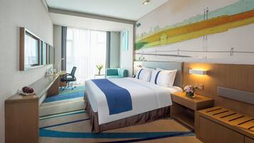 Quarto de hotel com cama, escrivaninha, cadeira, mesa de cabeceira e televisão fixada na parede.