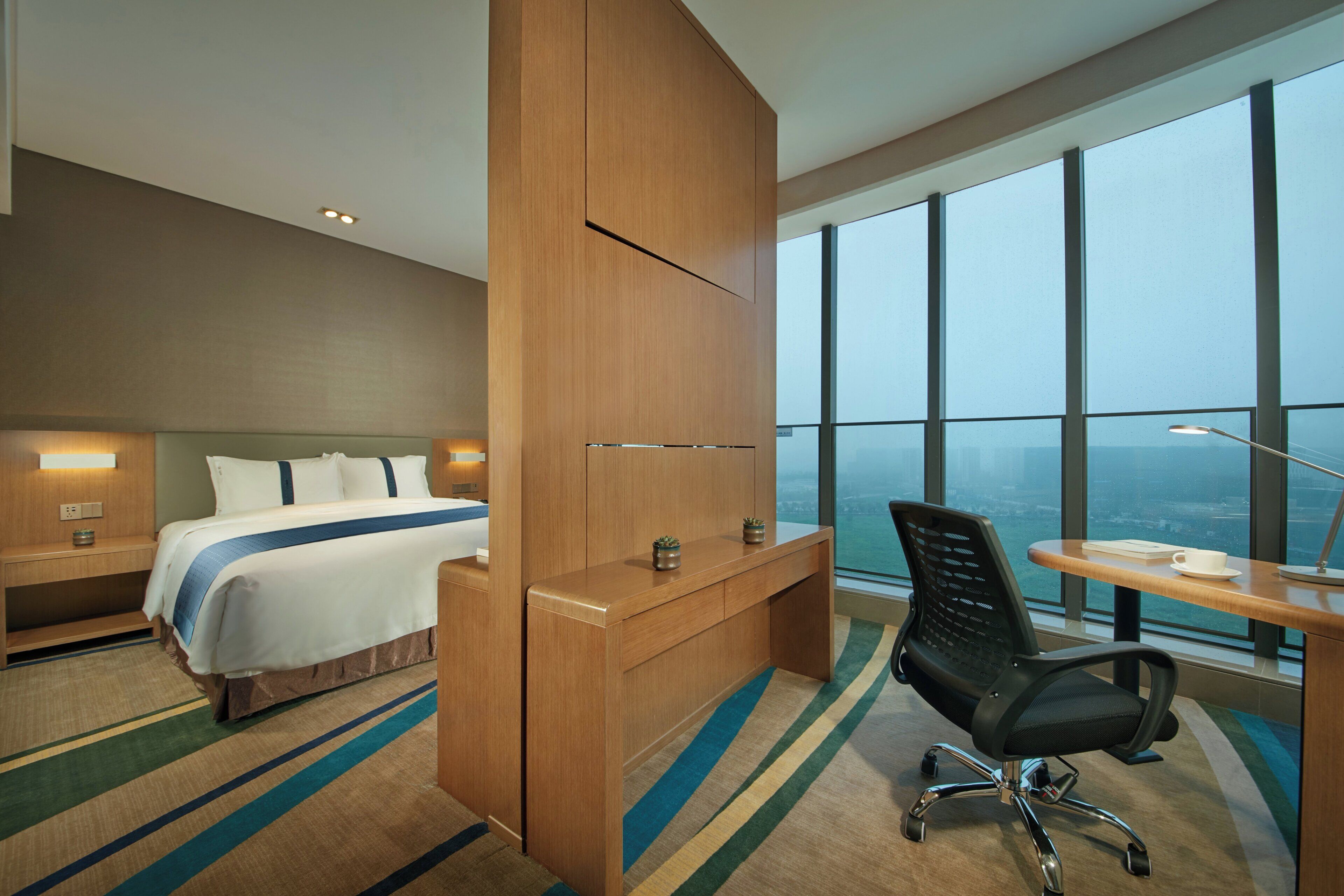 Quarto de hotel com cama, escrivaninha, cadeira e amplas janelas com vista para a cidade.