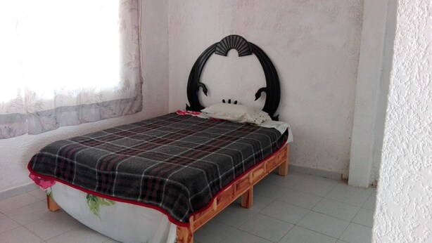 Quarto pequeno com uma cama de solteiro, estrutura de madeira, colcha xadrez e parede branca com cabeceira decorativa.