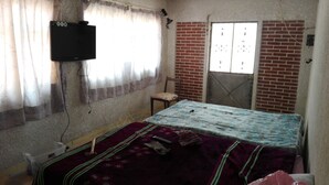 Um quarto com uma cama, uma televisão na parede, uma janela com vista para um muro de tijolos e uma porta.