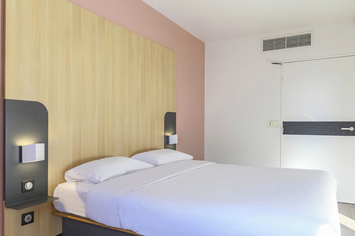 Uma cama bem arrumada num quarto de hotel, com cabeceira de madeira, abajures e uma porta branca.