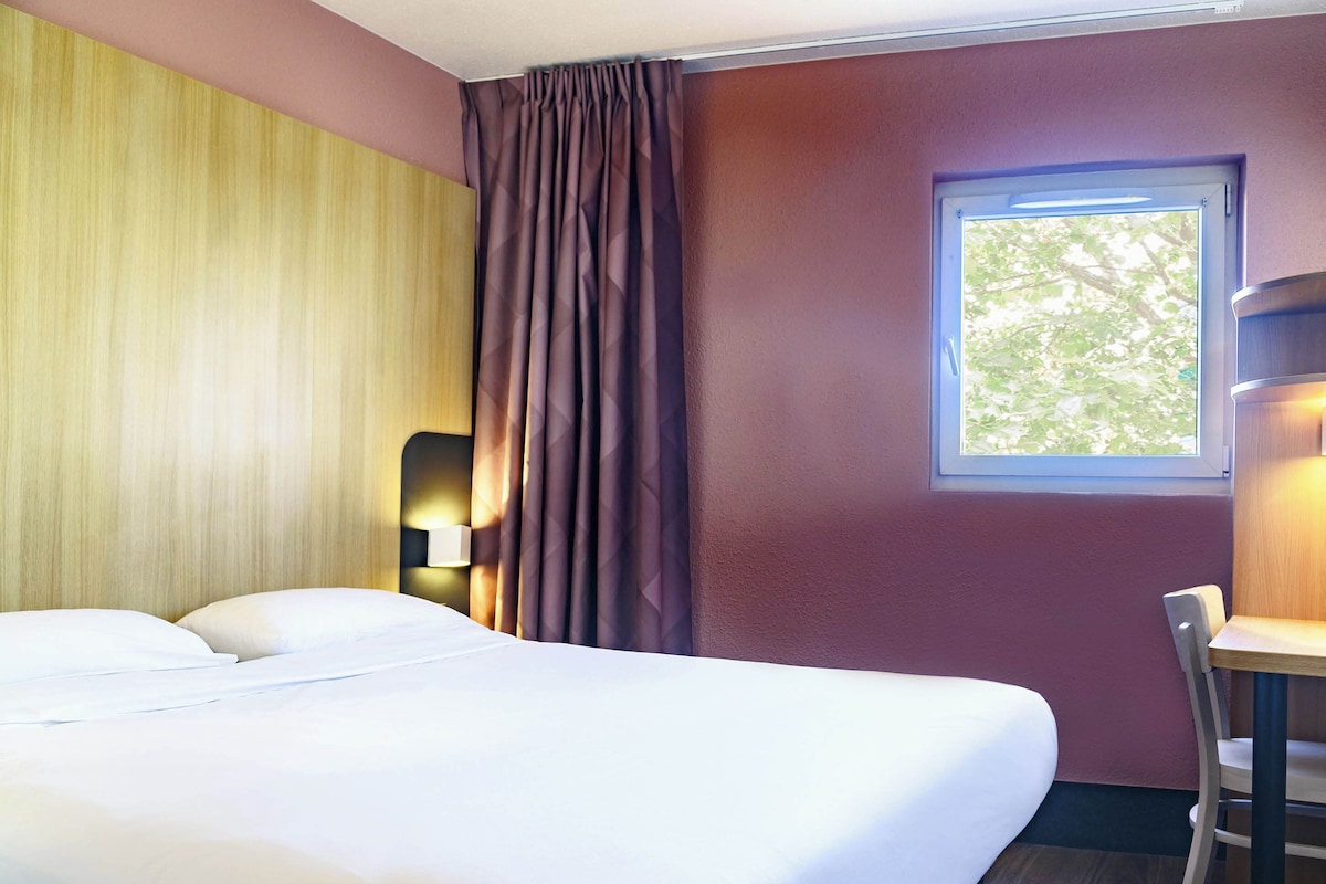 Quarto de hotel com cama, escrivaninha e janela com vista para árvores.