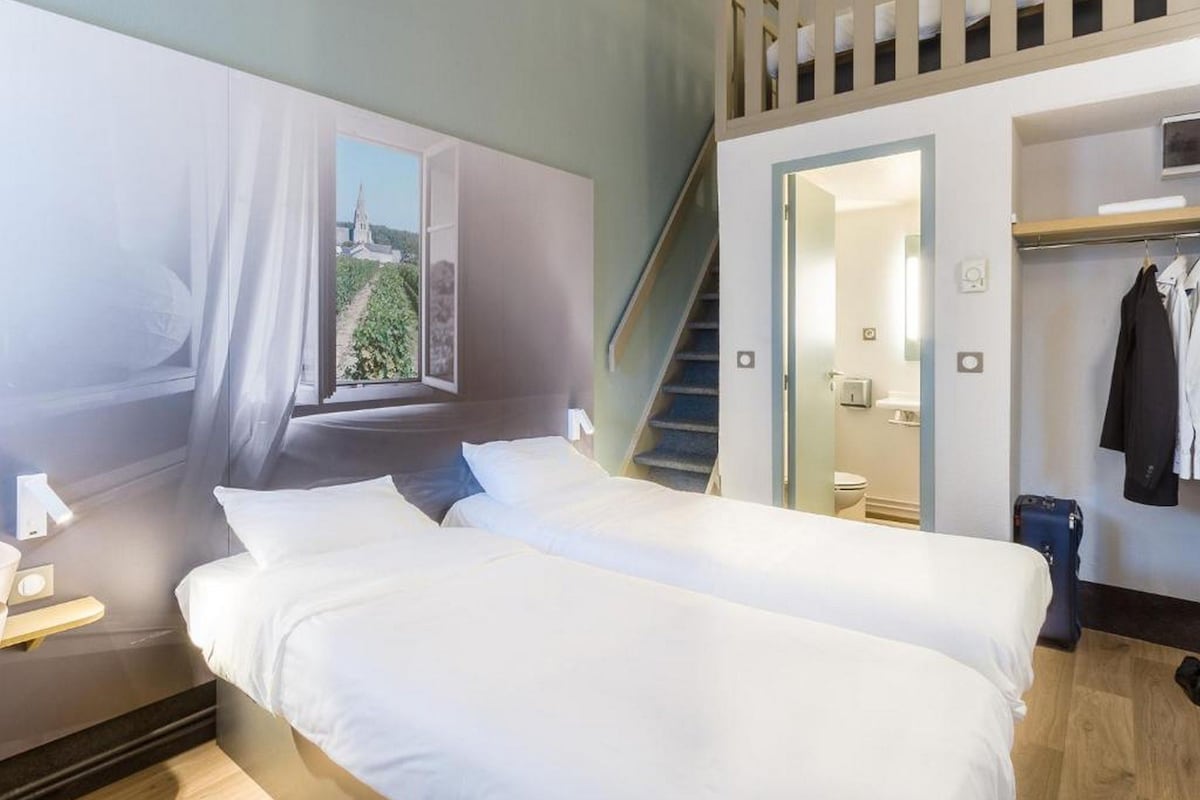 Quarto de hotel com duas camas, janela com vista, escada e porta-casacos.