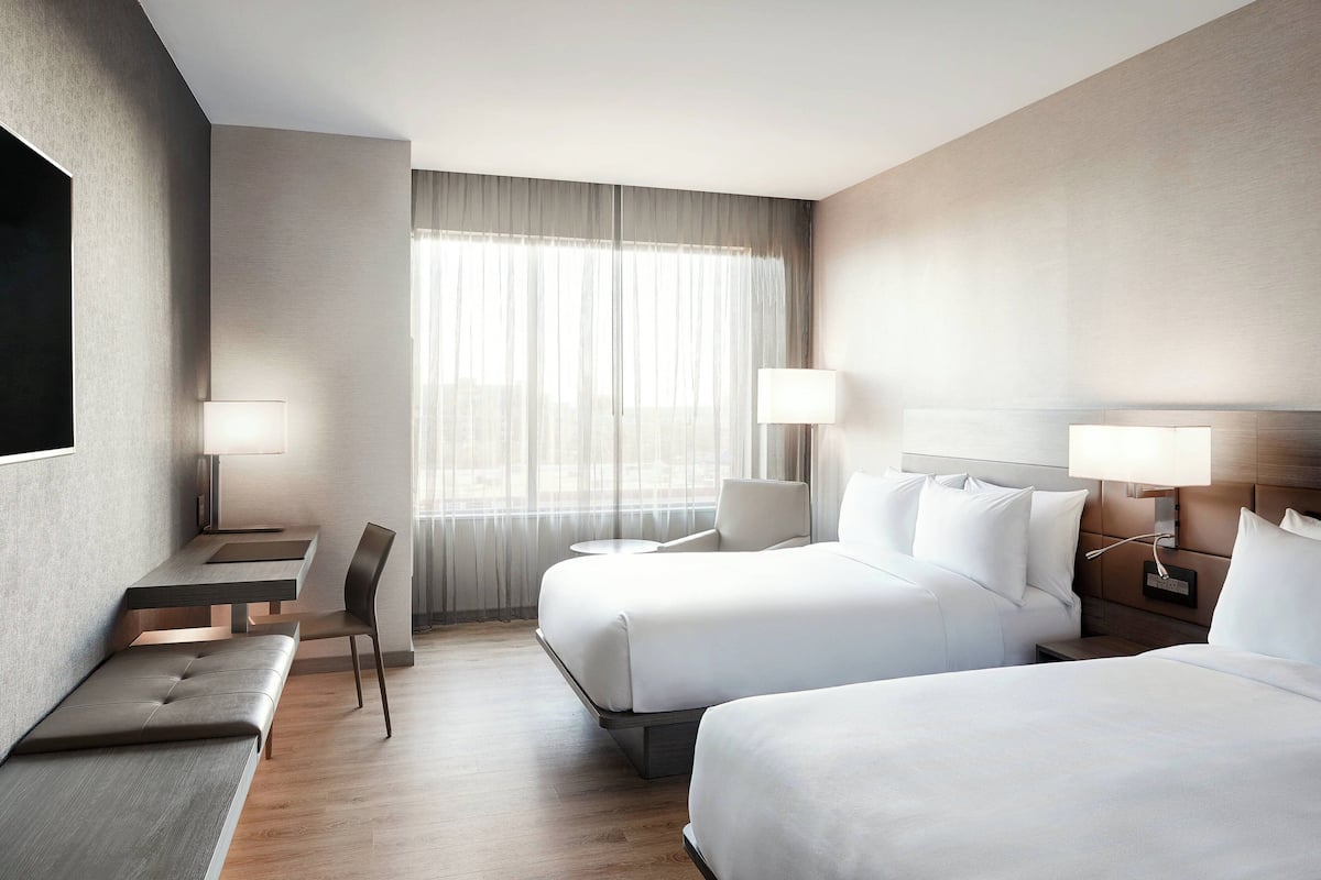 Quarto de hotel com duas camas, uma escrivaninha, uma cadeira e uma janela grande com cortinas translúcidas.