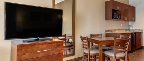 Quarto de hotel com televisão de tela plana, móveis de madeira, área de cozinha e um espelho grande.