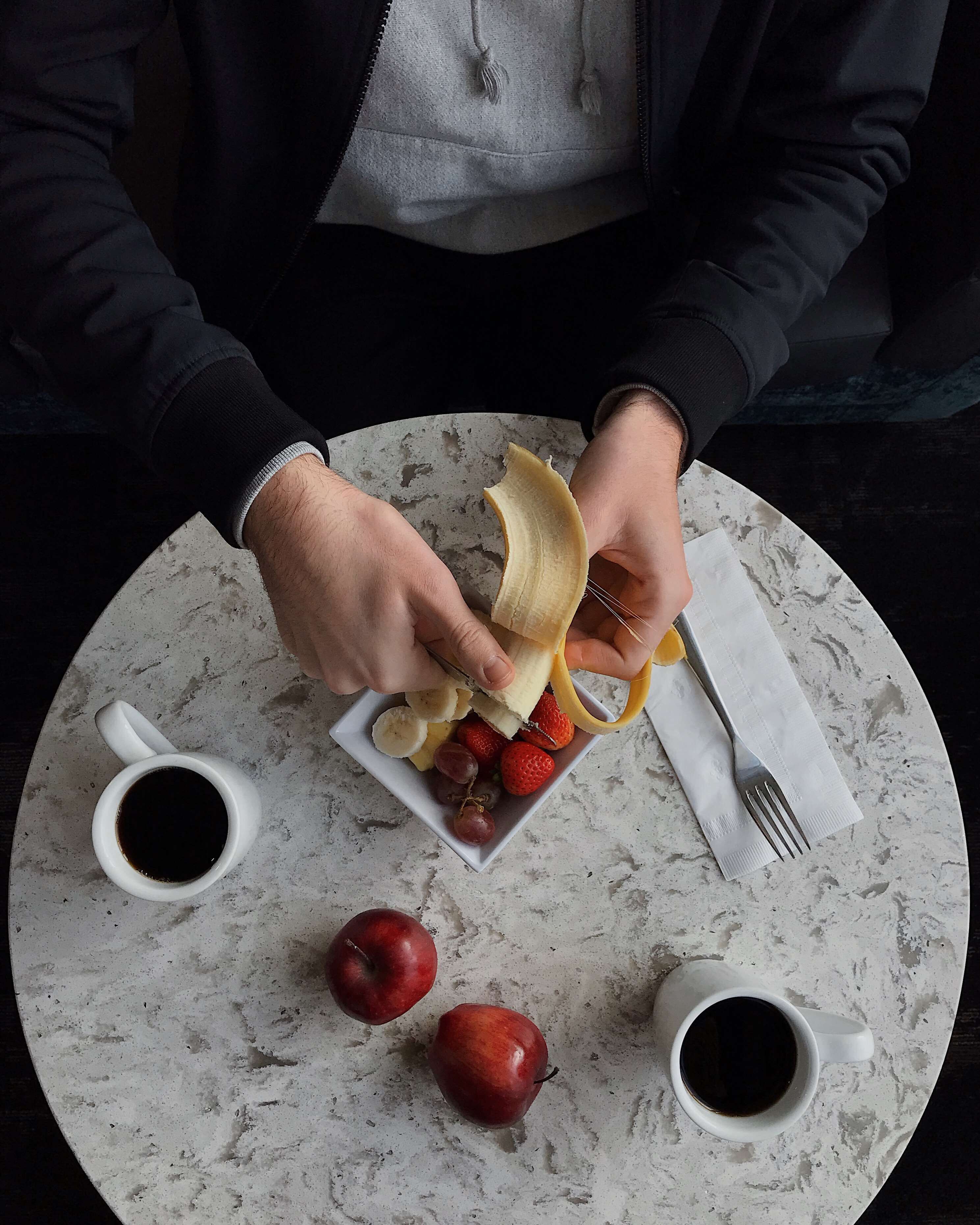 Uma pessoa descascando uma banana e cortando uma morango sobre uma mesa de mármore, com duas chávenas de café e duas maçãs.