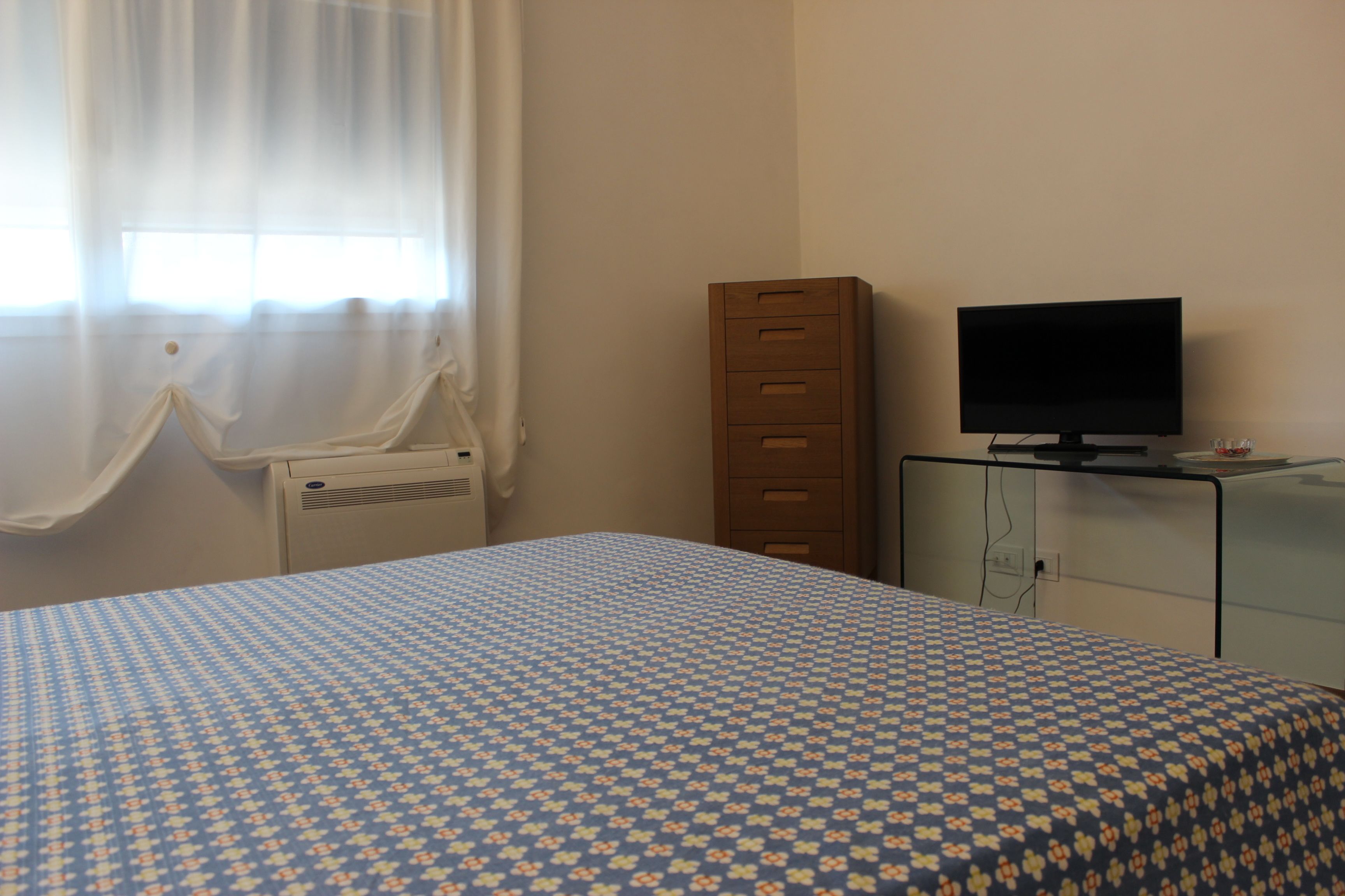 Quarto com cama, televisão em móvel, penteadeira e janela com cortinas leves.