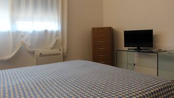 Quarto com cama, televisão em móvel, penteadeira e janela com cortinas leves.