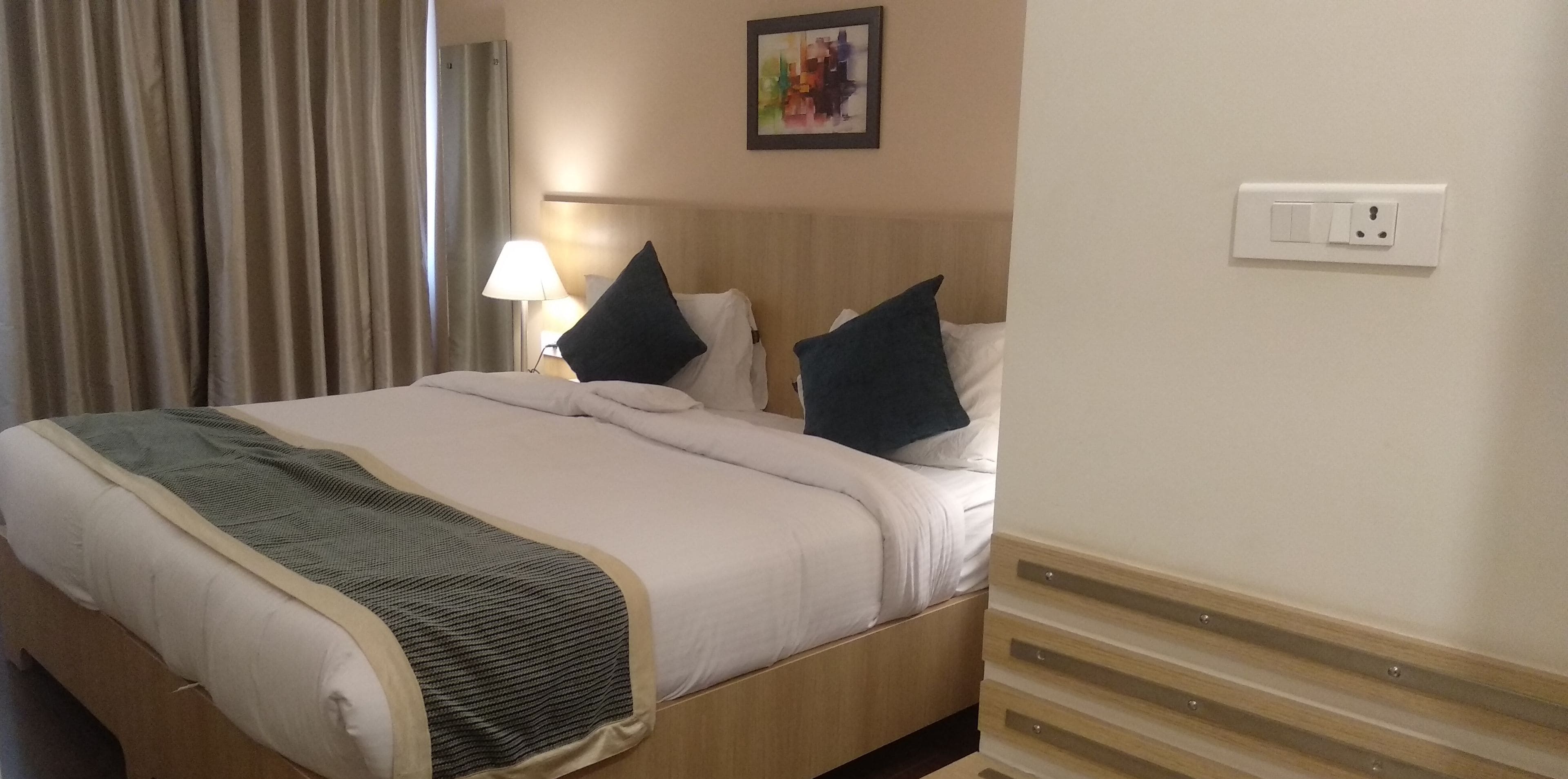 Quarto de hotel com cama, luminária, quadro na parede e tomada.