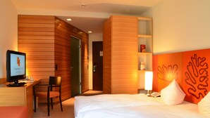 Quarto de hotel com uma cama, uma escrivaninha com computador, uma cadeira, uma televisão e um painel decorativo na parede.