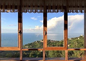 Vista pela janela com moldura de madeira e um valance decorativo, com vista para uma paisagem costeira com edifícios e vegetação.