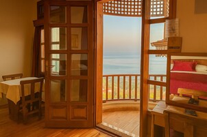 Quarto com porta de madeira que dá para uma varanda com vista para o mar. Há uma cama com roupa de cama vermelha e uma pequena mesa com cadeiras.