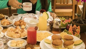 Mesa posta com diversos itens de café da manhã, incluindo pães, frutas e bebidas.