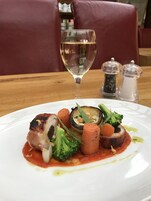 Um prato branco com uma refeição composta por carne, legumes e molho, um copo de vinho branco, e saleiros e pimenteiros sobre uma mesa de madeira.
