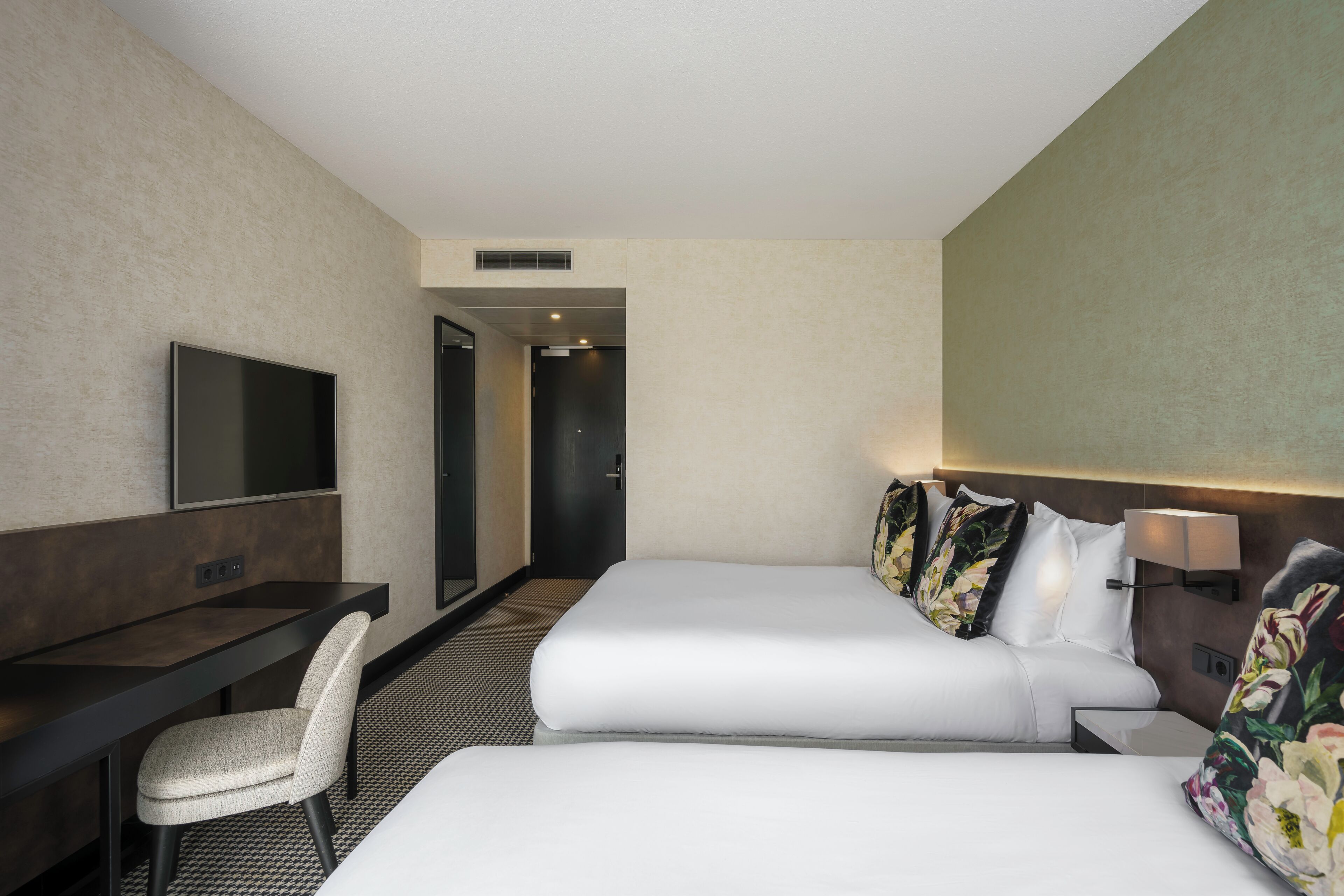 Quarto de hotel com duas camas, uma escrivaninha, uma cadeira, uma televisão e uma porta que dá para outro quarto.