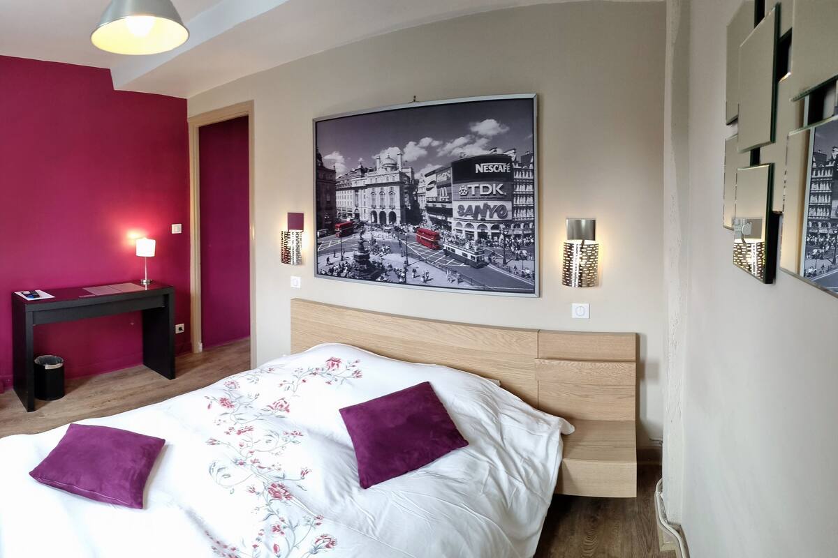 Quarto de hotel com cama, escrivaninha, cadeira, televisão na parede e um cartaz.