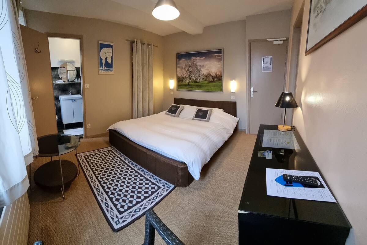 Quarto de hotel com cama, mesa de cabeceira, luminária e pequena escrivaninha com cadeira.