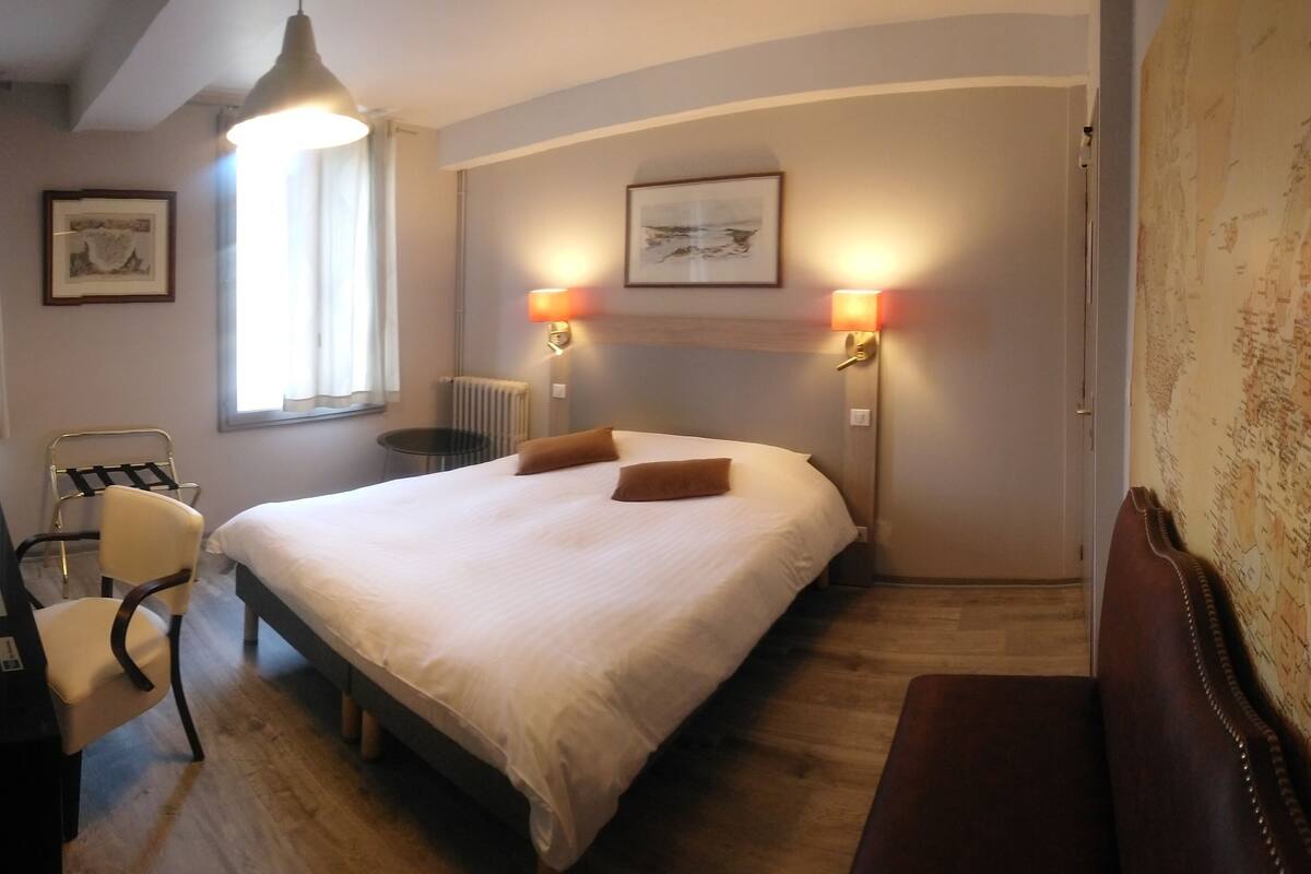 Quarto de hotel com cama, abajures, escrivaninha com cadeira, janela com cortinas e parede com mapa.