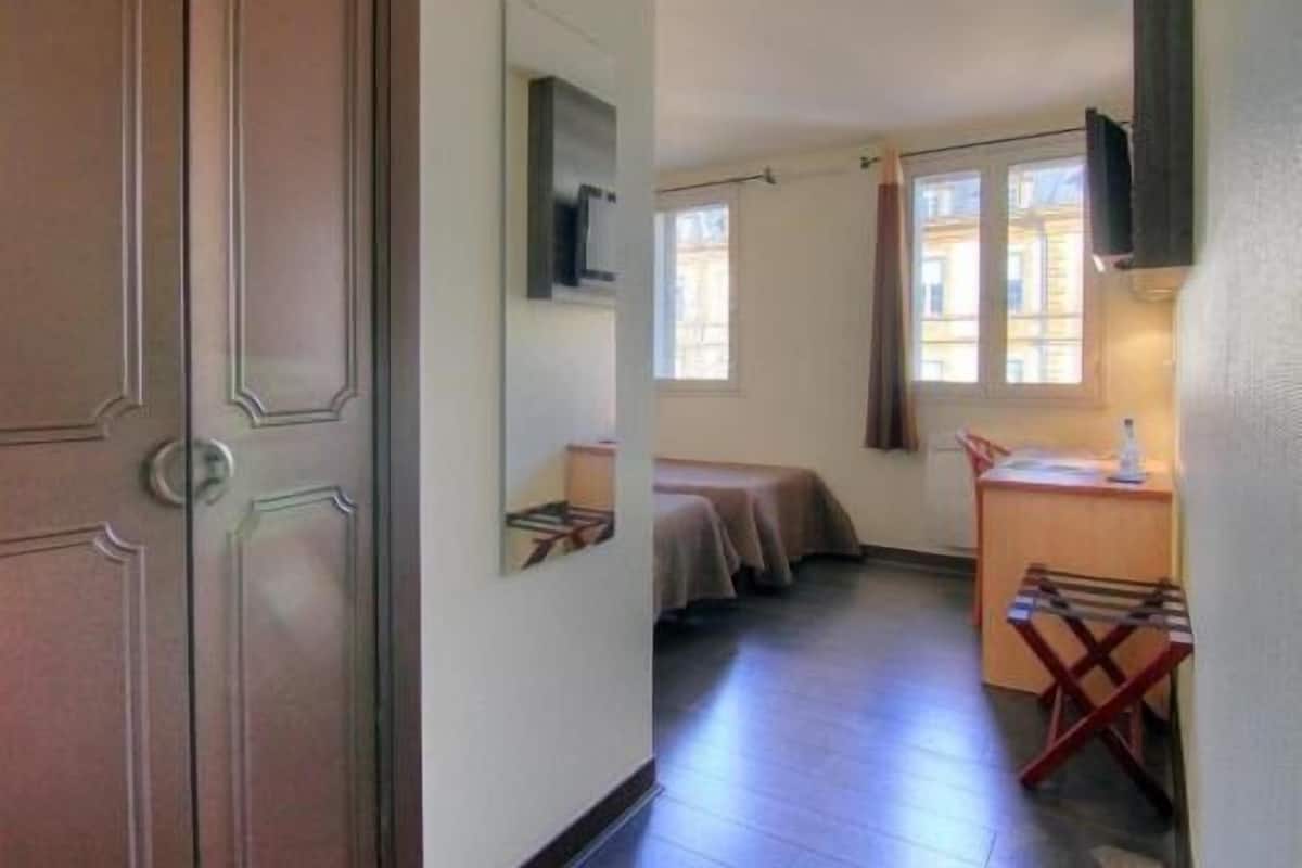 Quarto de hotel compacto com uma cama, uma pequena escrivaninha, uma cadeira e uma televisão.