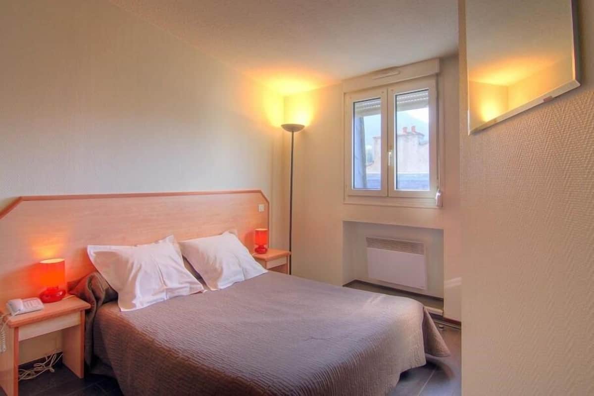Quarto de hotel com cama, luminárias de cabeceira, mesa de cabeceira, janela e espelho fixado na parede.