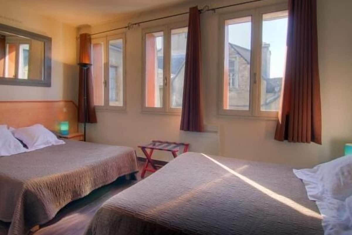 Quarto com duas camas, uma mesinha e janelas com cortinas.