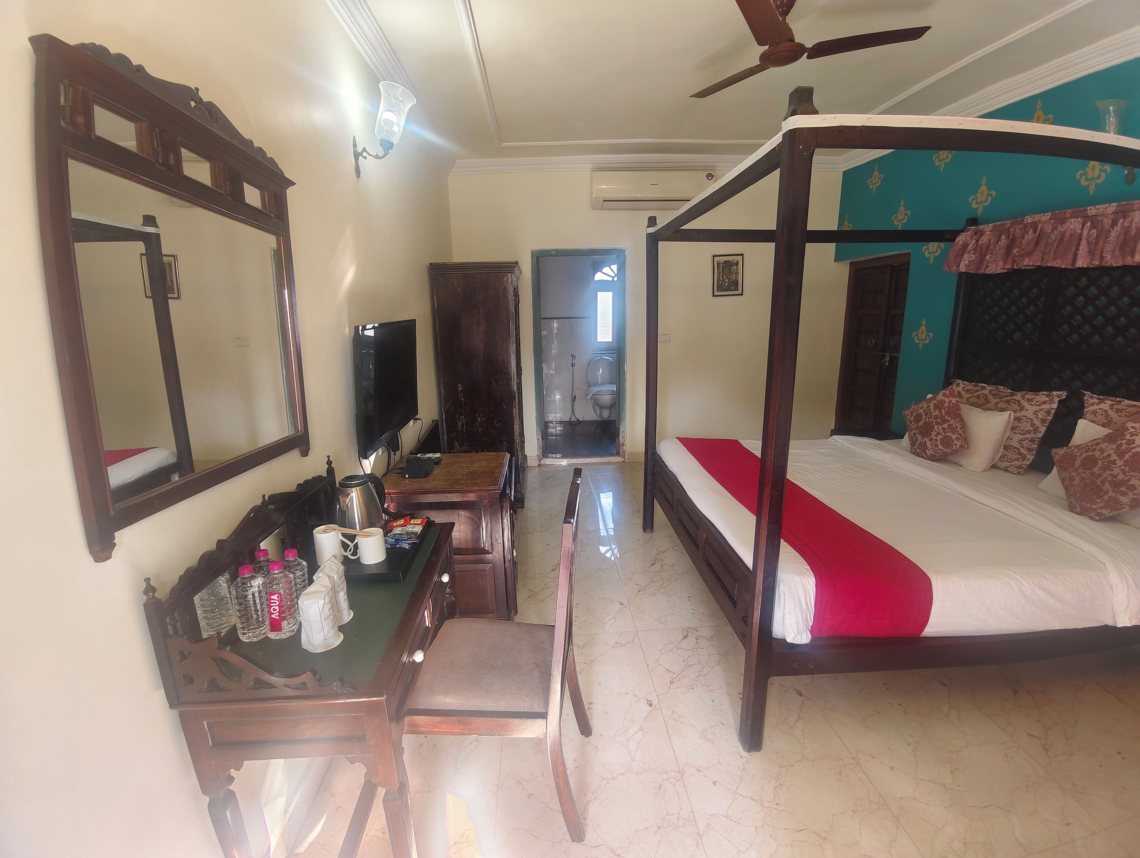 Quarto de hotel com cama de dossel, escrivaninha com cadeira, televisão e guarda-roupa.