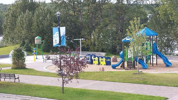 Parque infantil com escorregadores e estruturas de escalada, cercado por um espaço pavimentado e árvores.