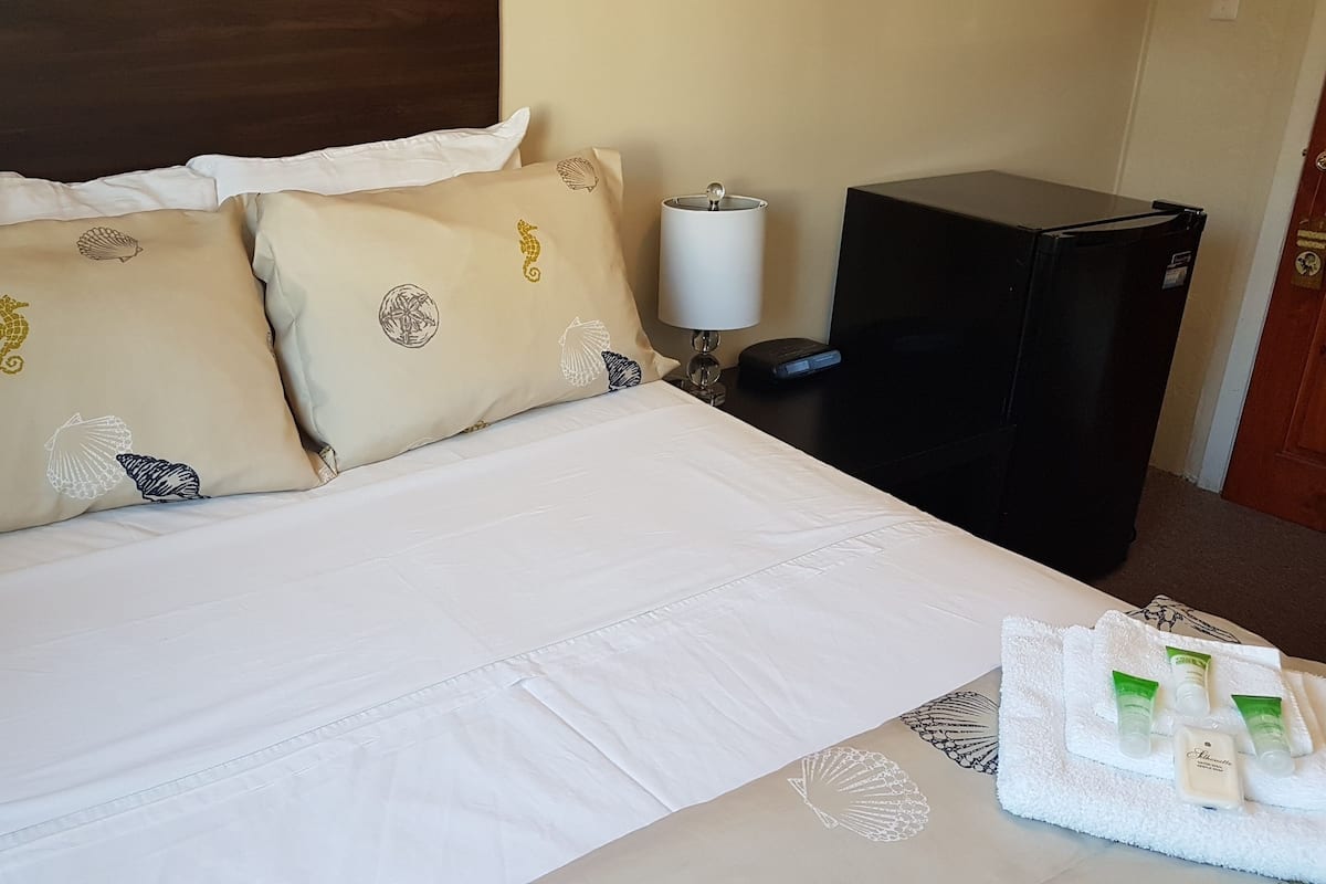 Quarto de hotel com cama, almofadas, luminária, geladeira preta e produtos de higiene em uma toalha.