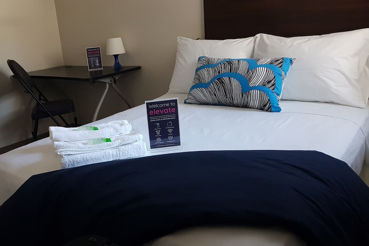 Quarto de hotel com cama, uma placa na cama, uma cadeira, uma mesa e um travesseiro.