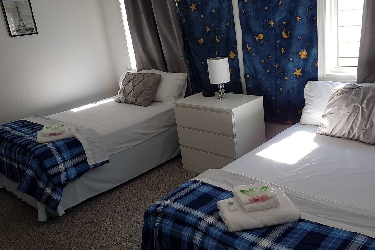 Quarto de hotel com duas camas, mesa de cabeceira, abajur e janela com cortinas.