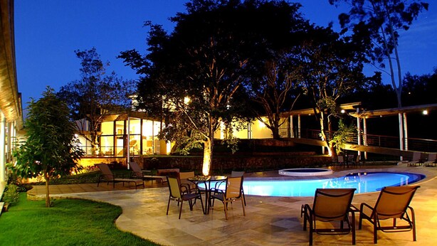 Uma área de piscina externa com mesa e cadeiras, cercada por um gramado bem cuidado e árvores.