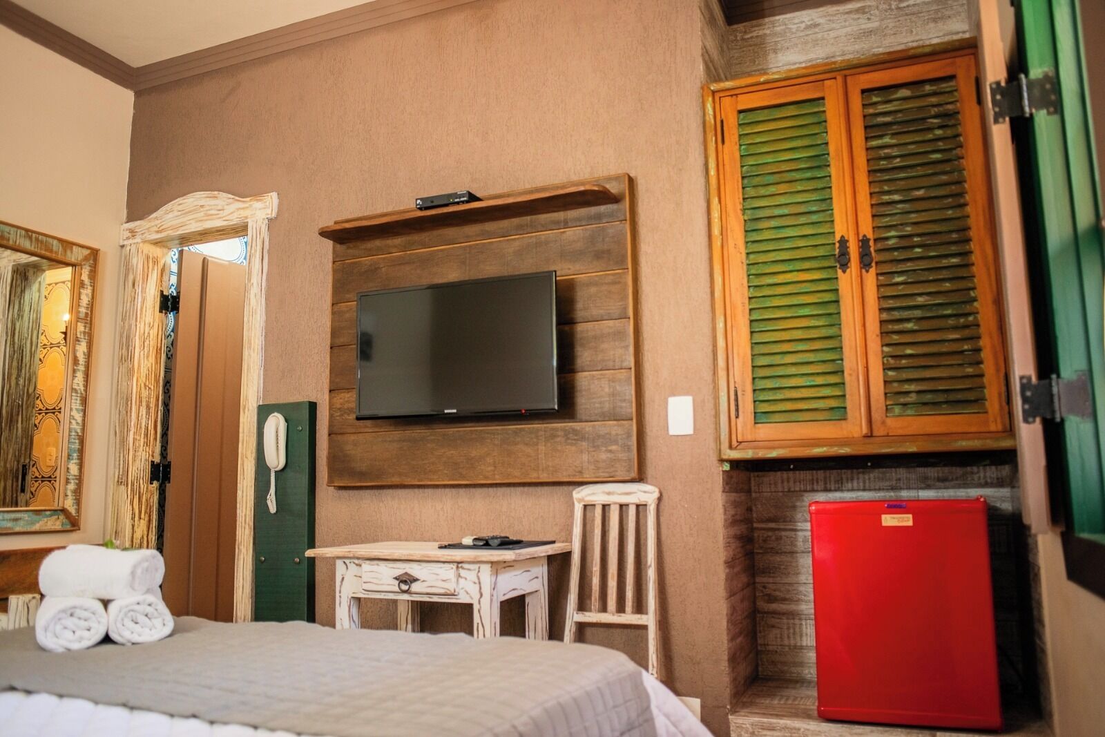 Quarto com cama, televisão na parede, mesinha, cadeira, guarda-roupa e frigorífico vermelho.