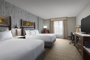 Quarto de hotel com duas camas, uma escrivaninha, uma cadeira, uma televisão e uma janela com cortinas.