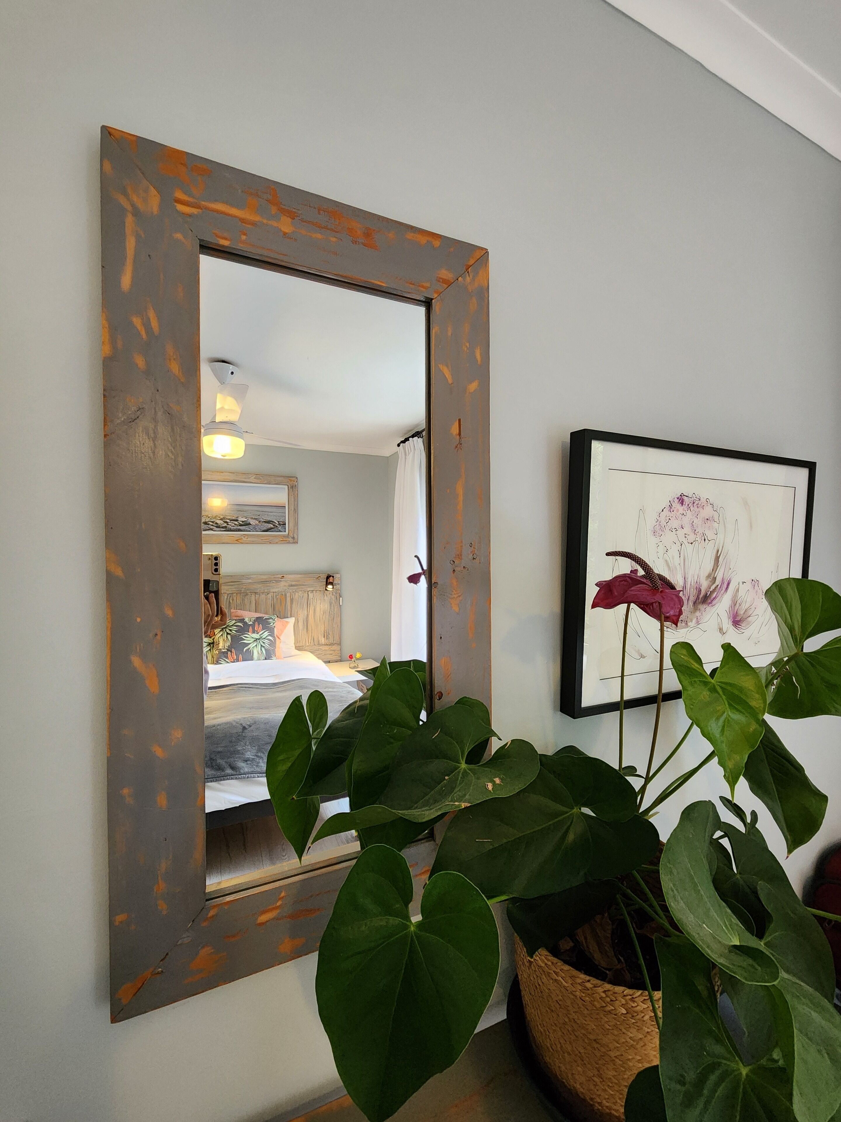Um espelho com moldura rústica reflete um quarto com uma cama, mesa de cabeceira e um quadro na parede.