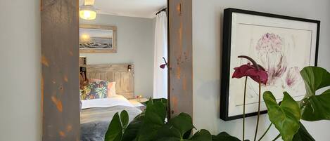 Um espelho com moldura rústica reflete um quarto com uma cama, mesa de cabeceira e um quadro na parede.