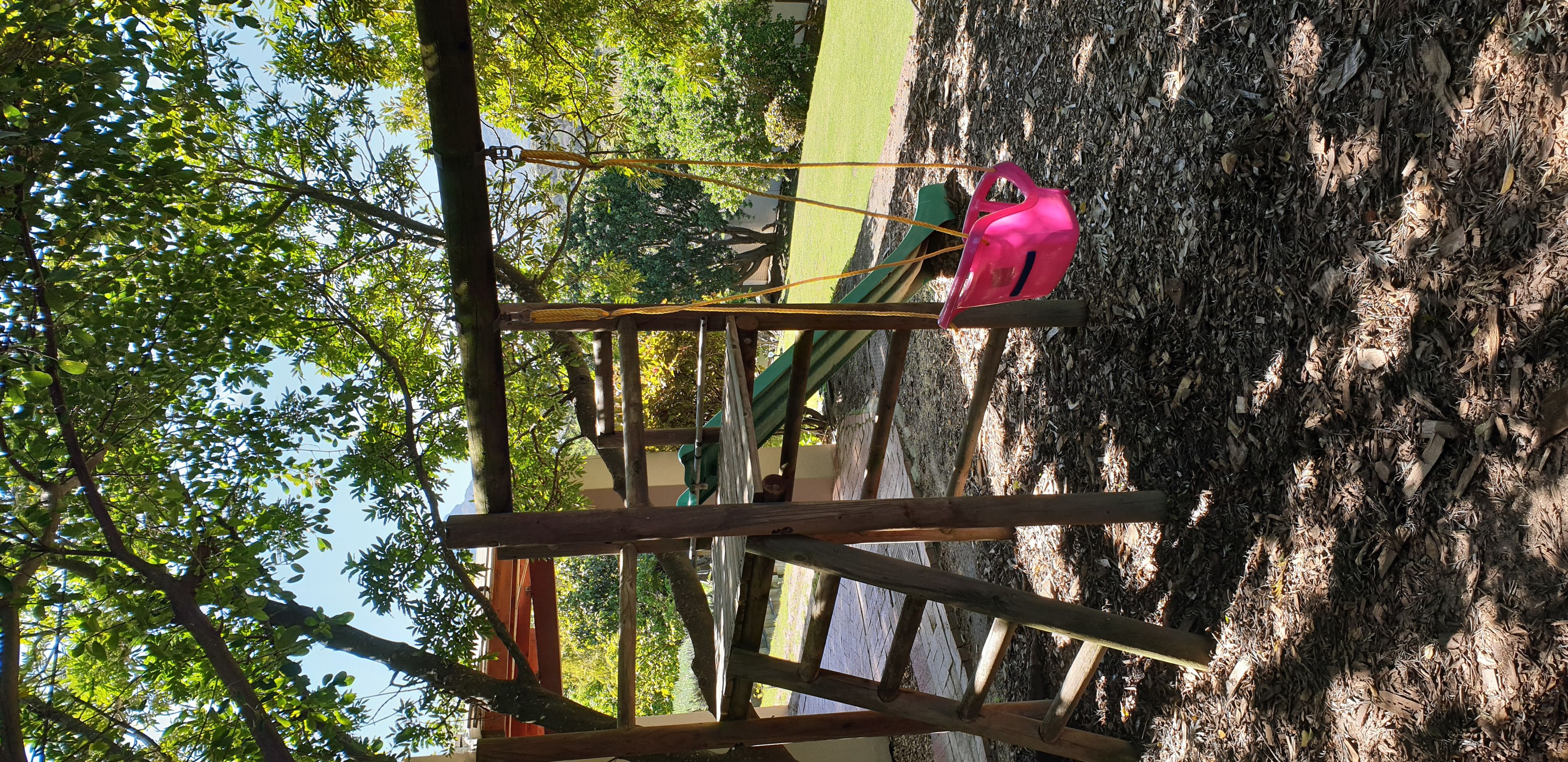 Um baloiço com um assento rosa e um assento verde, rodeado por árvores e um deck de madeira.