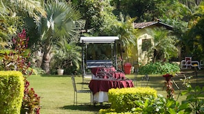 Um jardim com um gazebo, uma mesa com toalha vermelha e cadeiras.