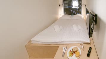 Cama bem arrumada com roupa de cama e toalhas brancas, mesa de cabeceira com prato de comida, xícara, talheres e controle remoto.